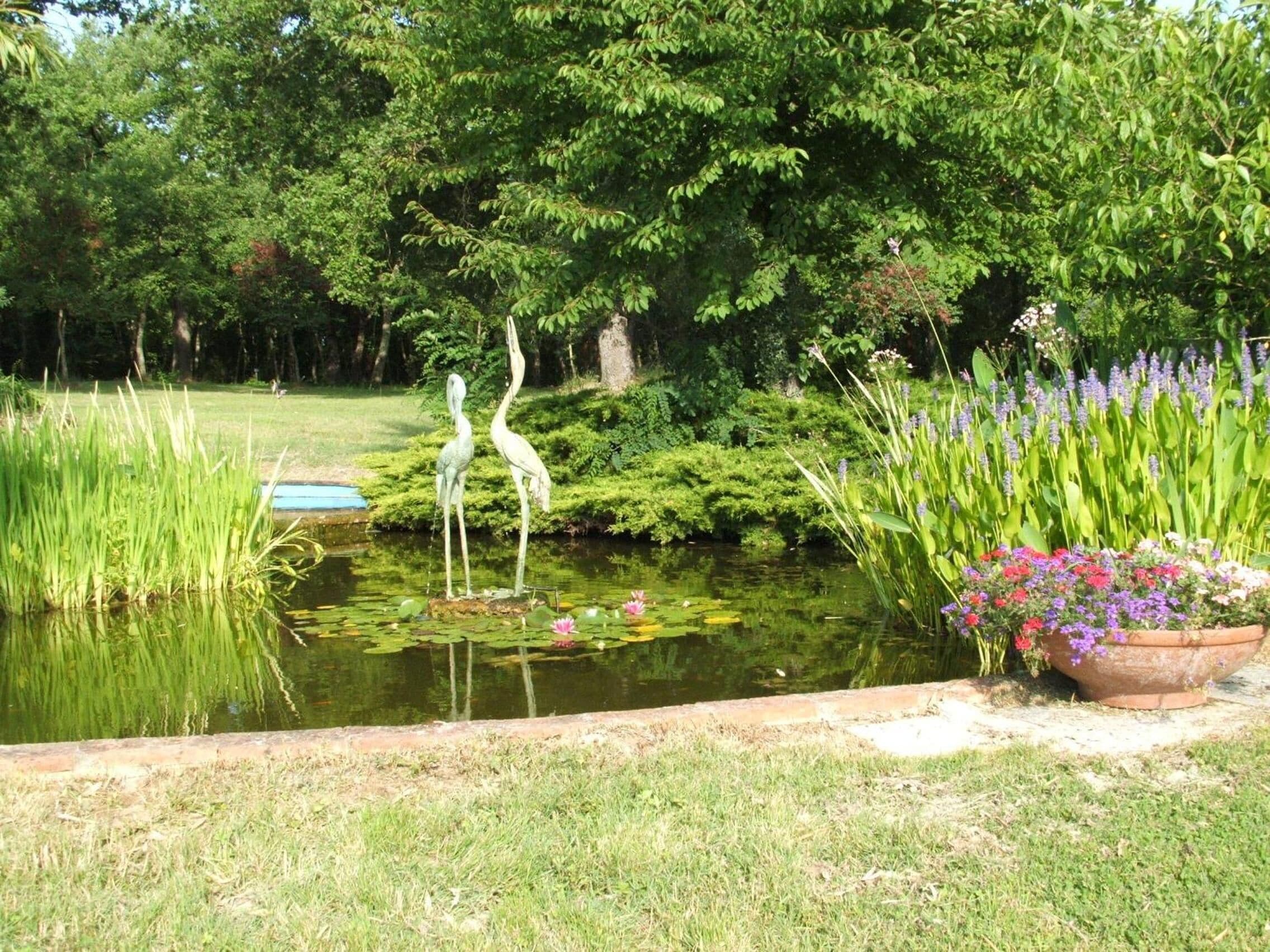 Um lago tranquilo com duas estátuas brancas de pássaros, rodeado por vegetação exuberante e diversas flores.