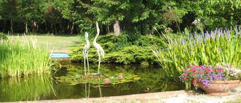 Um lago tranquilo com duas estátuas brancas de pássaros, rodeado por vegetação exuberante e diversas flores.