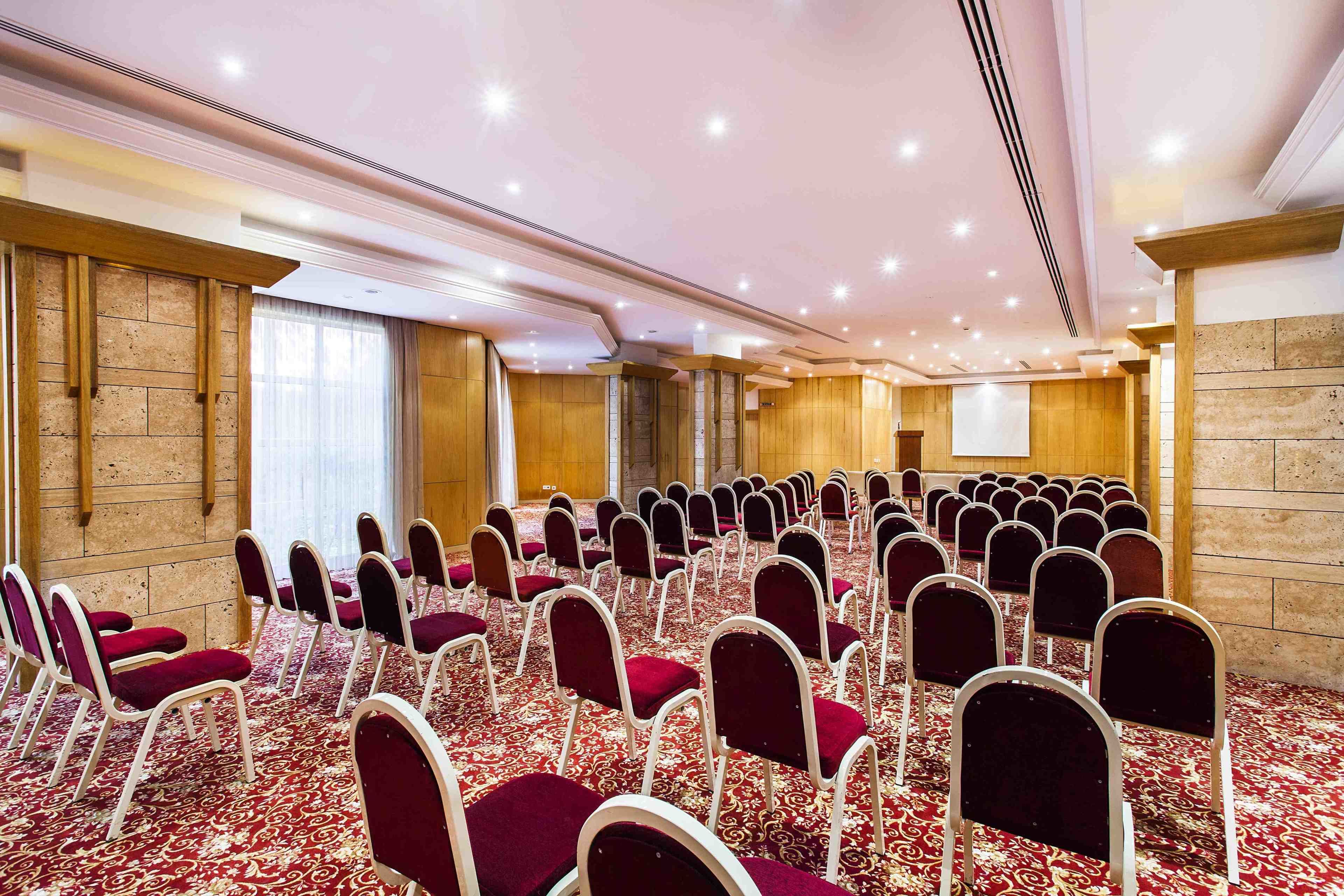 Uma sala de conferências com fileiras de cadeiras, piso carpete e tela de projeção.