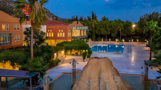 Um hotel com piscina, palmeiras e montanhas ao fundo.