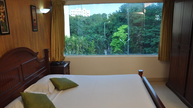 Quarto de hotel com cama grande, cabeceira de madeira, duas almofadas verdes, mesa de cabeceira, janela com cortinas e vista para árvores e edifícios.
