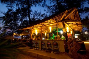 Um bar rústico ao ar livre com vigas de madeira, telhado de palha e vários bancos.
