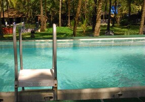 Piscina com escada de metal, cercada por vegetação exuberante e uma construção com telhado de palha ao fundo.