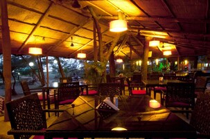 Um restaurante ao ar livre com mesas e cadeiras de madeira, telhado de palha e luminárias pendentes.