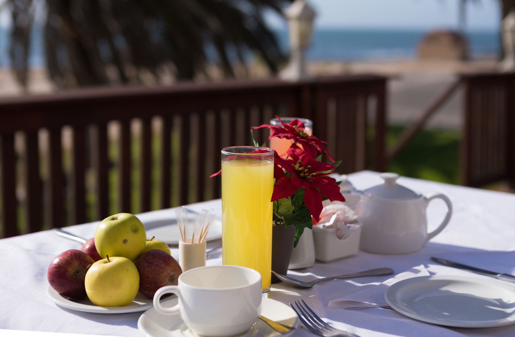 Mesa de café da manhã com frutas, um copo de suco de laranja, uma xícara, um bule e vista para o oceano.