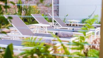 Área de lounge ao ar livre, iluminada pelo sol, com cadeiras brancas e pequenas mesas redondas, cercada por vegetação e um muro de tijolos.