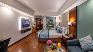Quarto de hotel com cama, sofá, televisão, mesa de jantar com comida e banheiro visível por uma porta aberta.