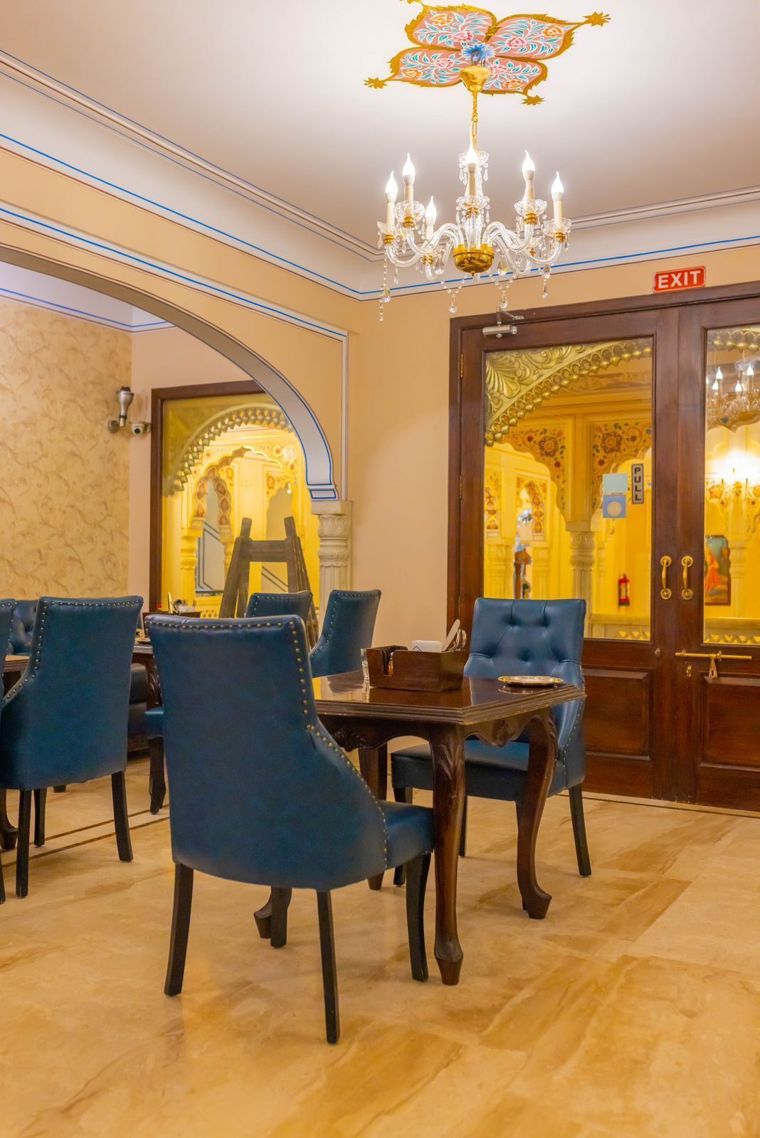 Uma sala de jantar com cadeiras estofadas em azul, uma mesa de madeira e um lustre. Um arco dá acesso a um espaço com uma porta dourada e uma placa de SAÍDA.