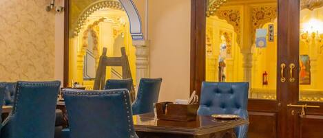 Uma sala de jantar com cadeiras estofadas em azul, uma mesa de madeira e um lustre. Um arco dá acesso a um espaço com uma porta dourada e uma placa de SAÍDA.