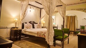 Uma cama com dossel e cortinas florais, uma escrivaninha com luminária e uma poltrona estofada verde.