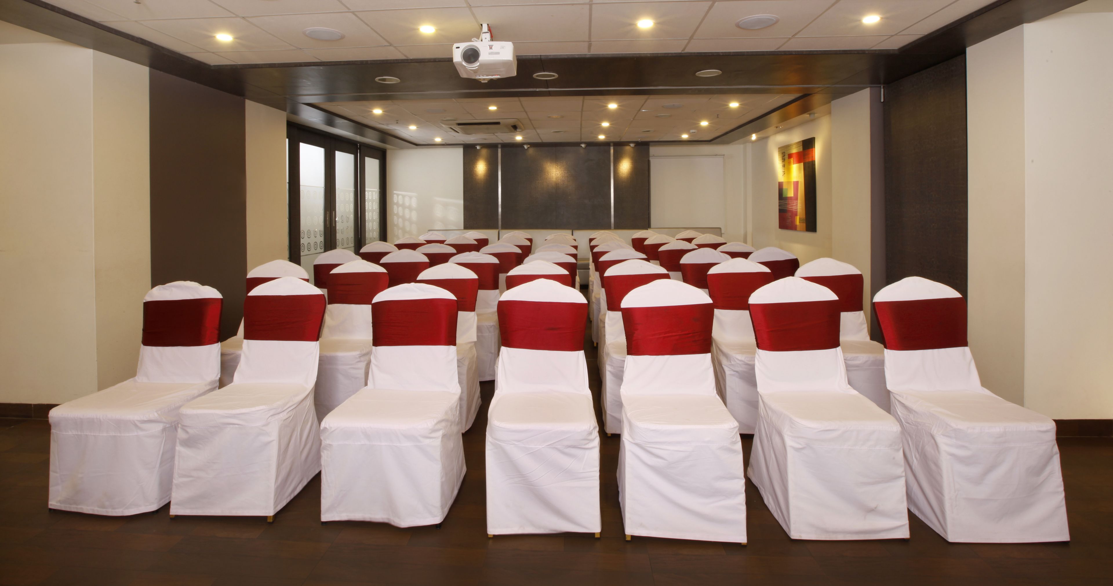 Sala de conferências com fileiras de cadeiras brancas com fitas vermelhas, uma tela de projetor e uma janela grande.