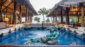 Uma área de piscina com uma estátua de um casal, estruturas de madeira com telhados de palha e vista para o mar.
