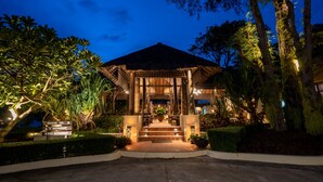 Um edifício bem iluminado, em estilo tropical, com telhado de palha, rodeado por vegetação exuberante e entrada pavimentada.
