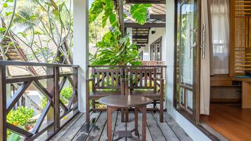 Um deck de madeira com mesa e cadeiras, rodeado por vegetação exuberante e vista para um edifício com grandes janelas.
