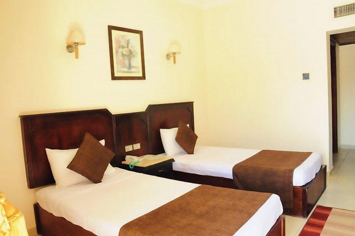 Quarto de hotel com duas camas, cada uma com cabeceira de madeira, roupa de cama branca e mantas marrons. Há duas luminárias fixadas na parede e um quadro emoldurado.