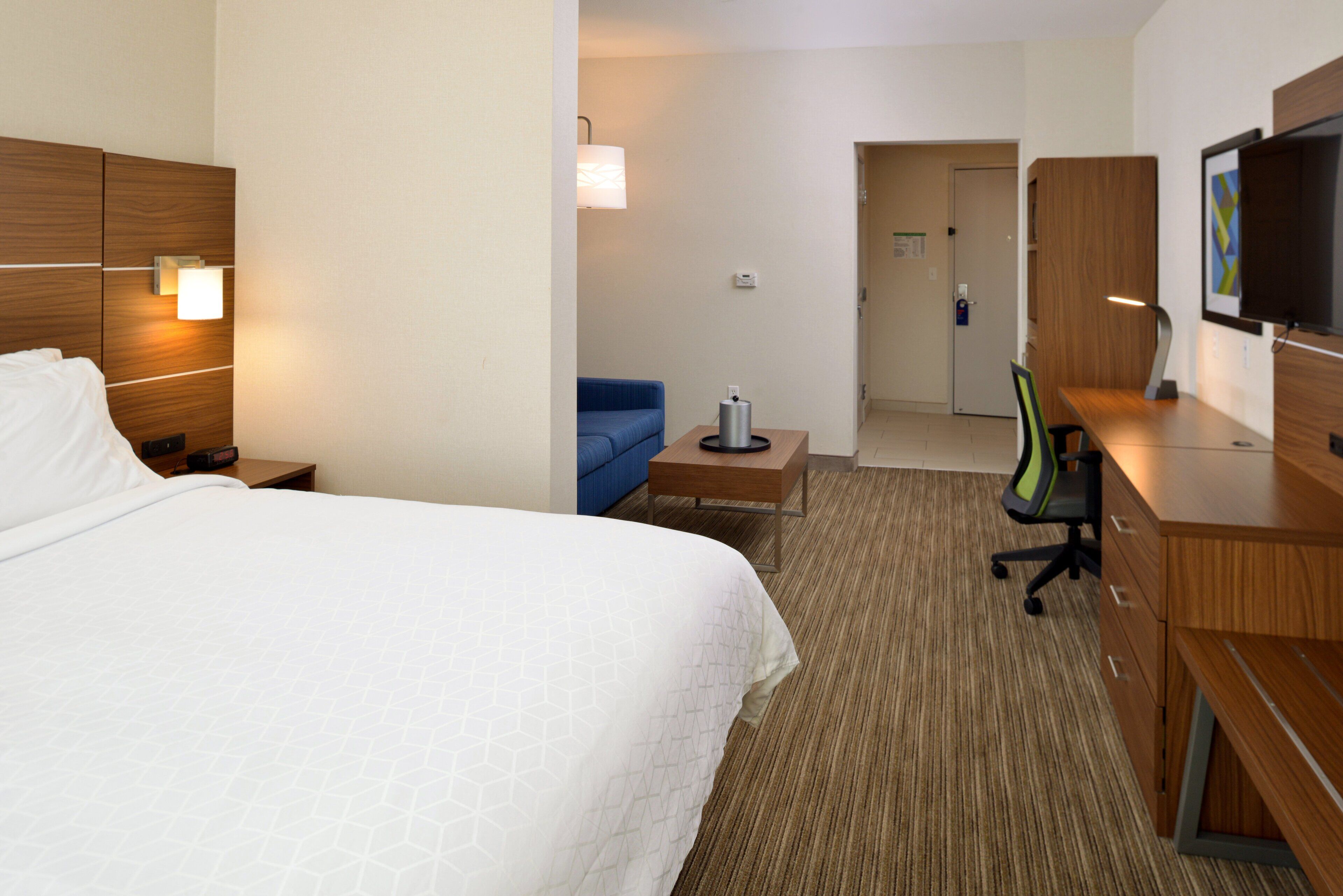 Quarto de hotel com uma cama grande, uma escrivaninha com cadeira, uma mesinha de cabeceira com abajur e uma televisão sobre um suporte.