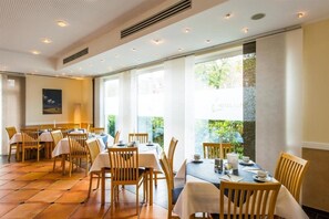 Espaço de refeições de hotel com várias mesas e cadeiras, janelas amplas e vista para o verde lá fora.