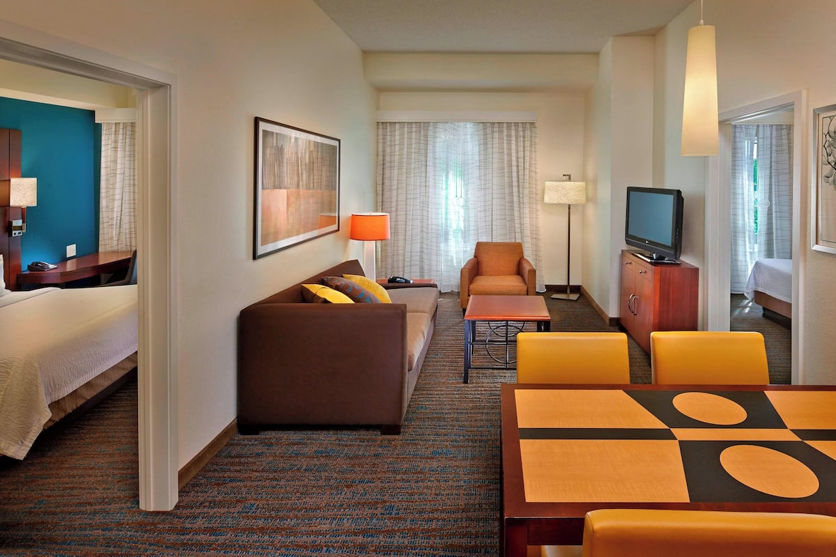 Quarto de hotel com cama, sofá, mesa de jantar com cadeiras, televisão e escrivaninha com luminária.