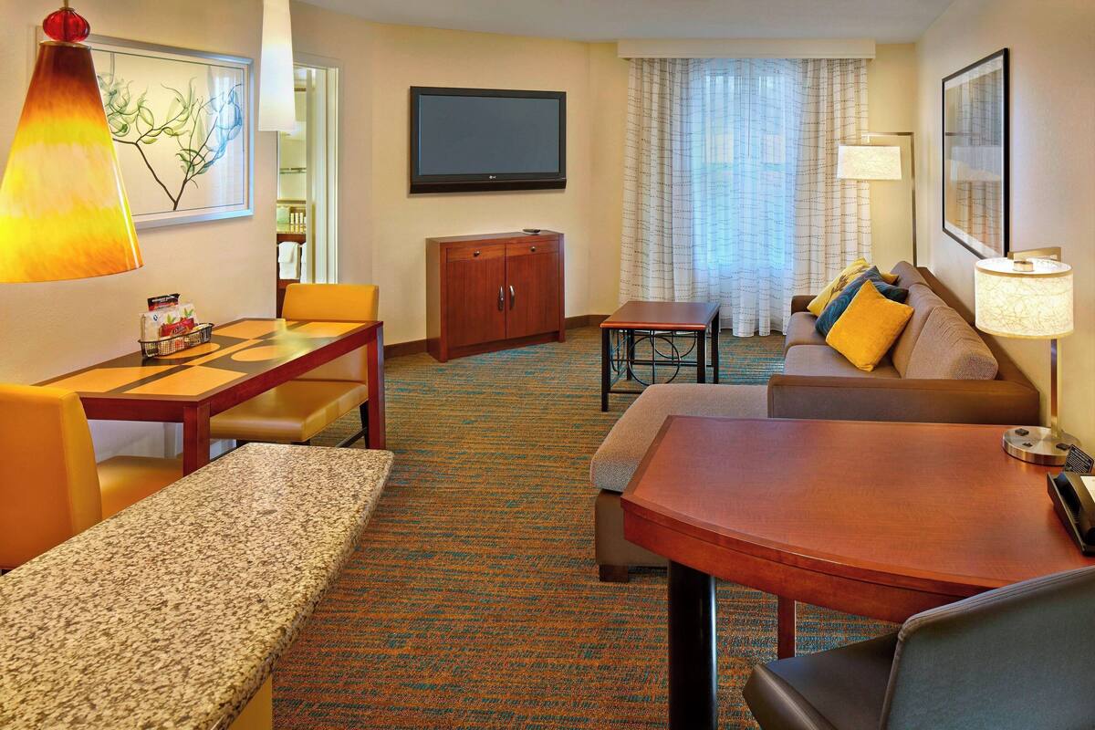 Quarto de hotel com mesa de jantar, sofá e televisão fixada na parede.