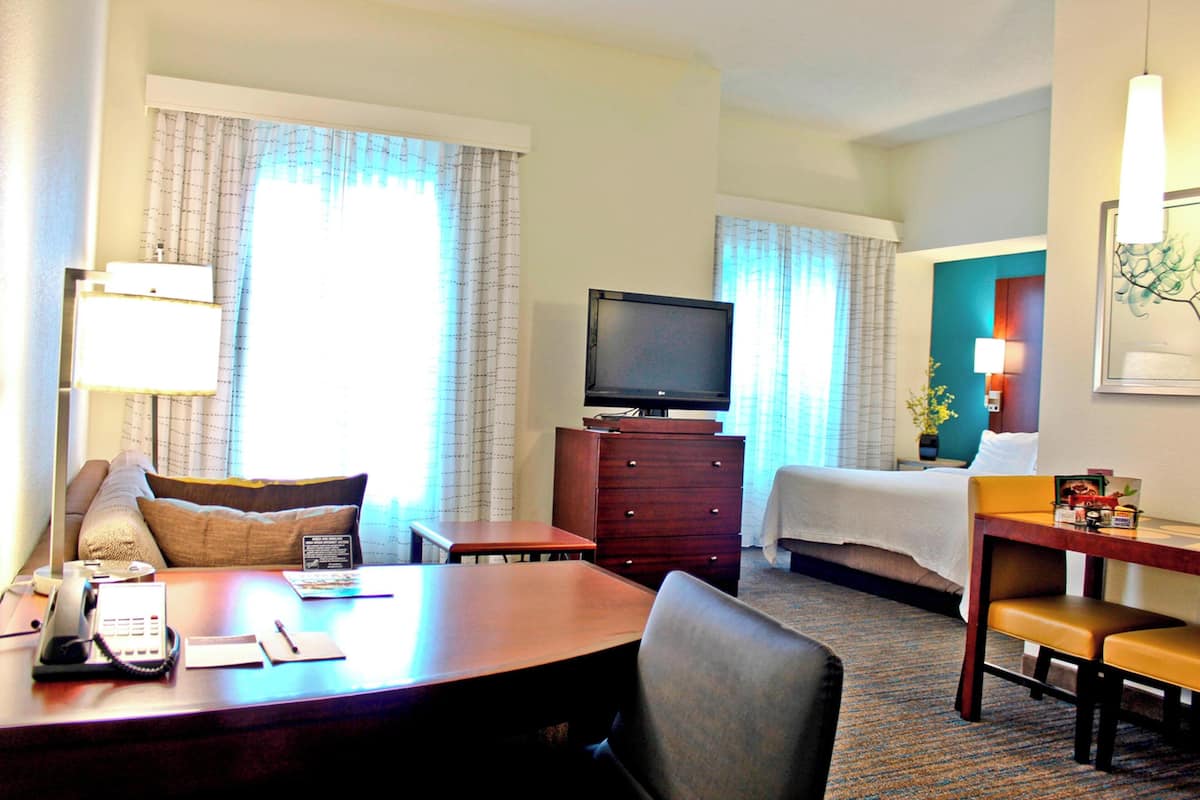 Quarto de hotel com uma cama, uma escrivaninha com telefone e papéis, uma televisão e uma cadeira.