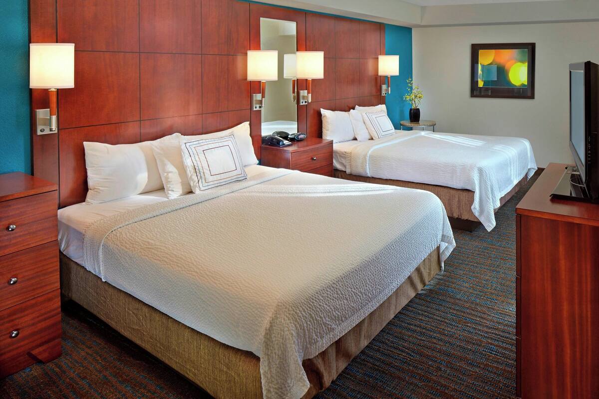 Quarto de hotel com duas camas, cabeceira de madeira, televisão de tela plana, escrivaninha e espelho.