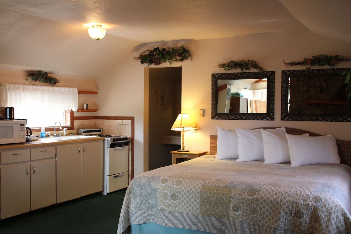 Quarto de hotel compacto com cozinha compacta, cama com várias almofadas, luminária de cabeceira e espelho na parede.