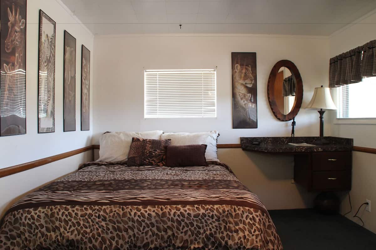 Um quarto com uma cama, janela com persianas, espelho, abajur e uma obra de arte na parede.