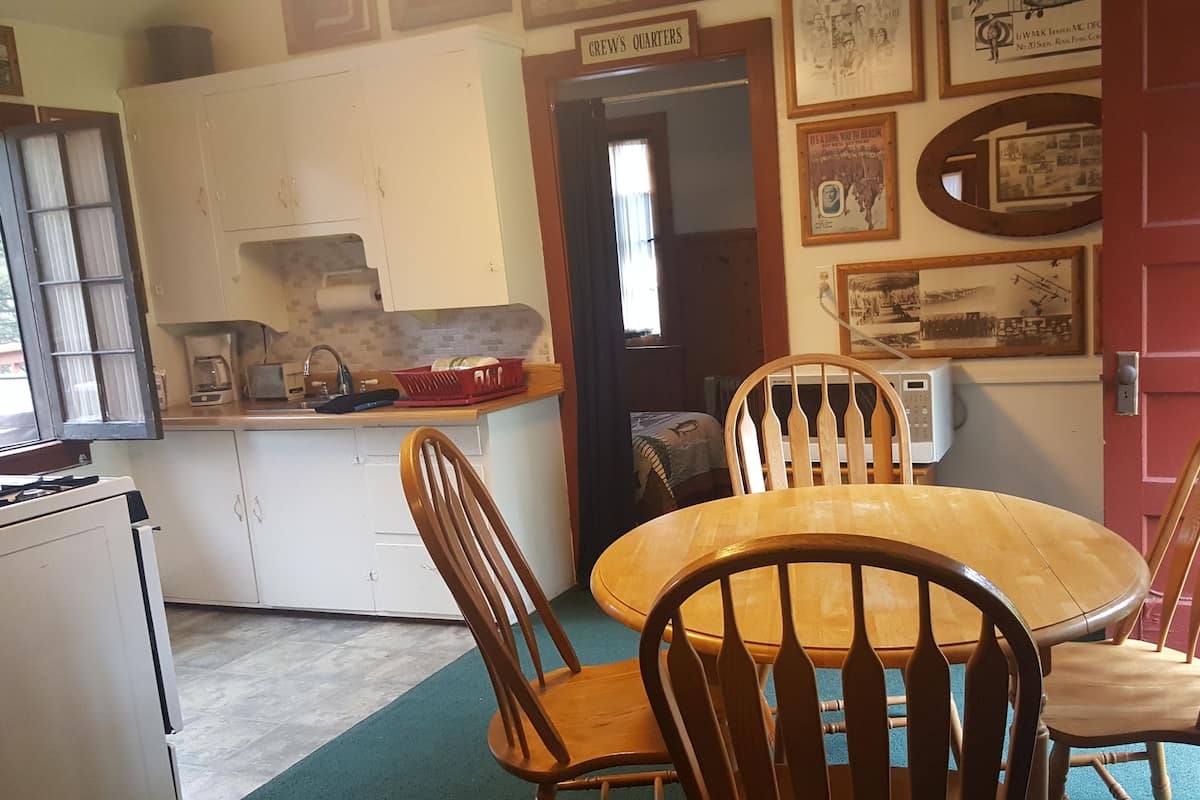 Uma sala de jantar com mesa e cadeiras de madeira, uma cozinha com pia e armários, e uma porta vermelha.