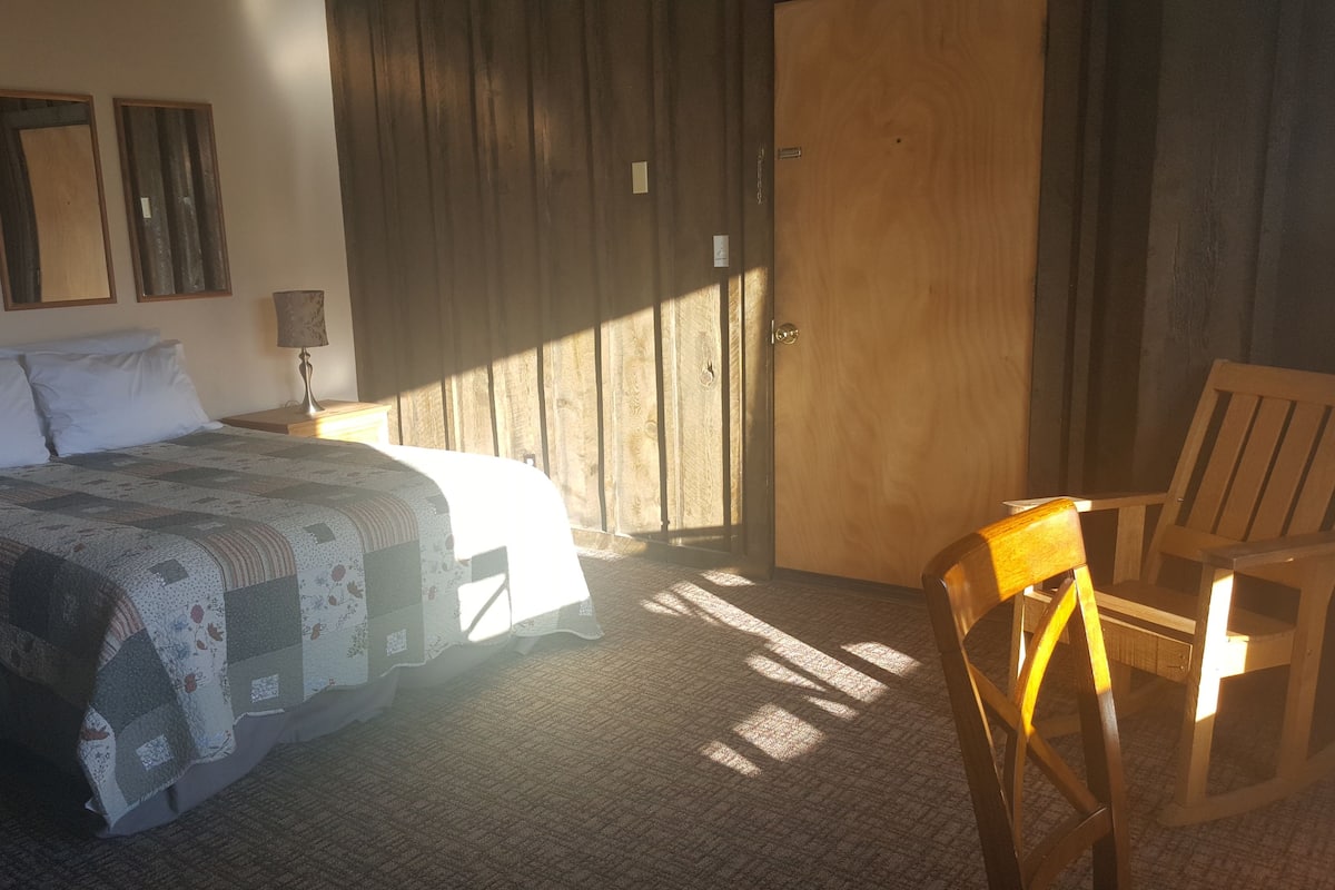 Quarto com uma cama, porta de madeira, uma cadeira e mesa de cabeceira com abajur.