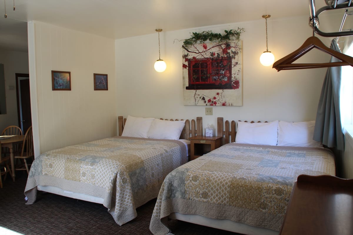 Quarto de hotel com duas camas, cabeceira de madeira, uma mesinha e uma cadeira. Há um quadro na parede e um lustre.