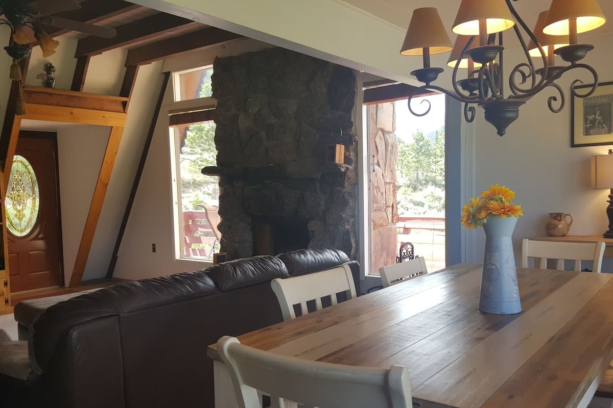 Uma sala de jantar com mesa de madeira, cadeiras brancas, lareira de pedra e um candelabro.