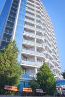 Um edifício alto com várias varandas, fachada de vidro e uma pequena área externa com bancos.