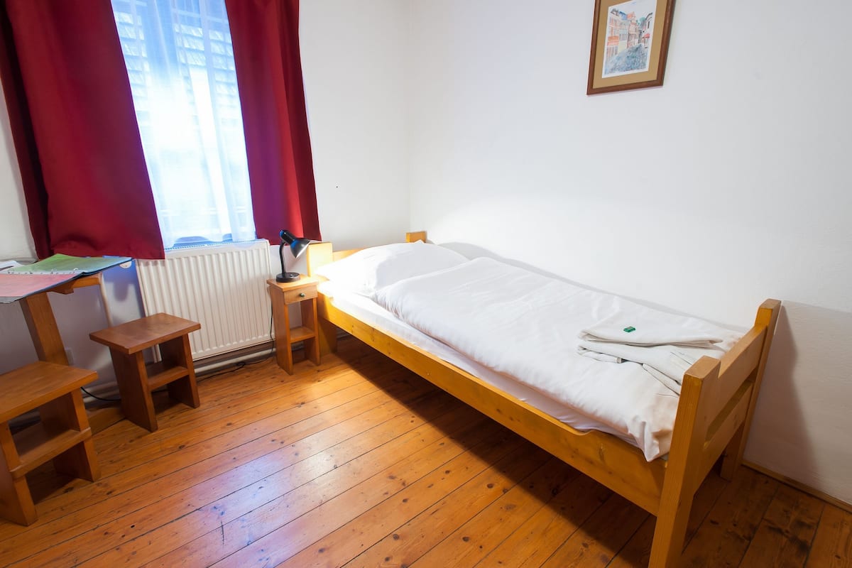 Quarto pequeno com uma cama de solteiro, soalho de madeira, escrivaninha, cadeira e radiador.