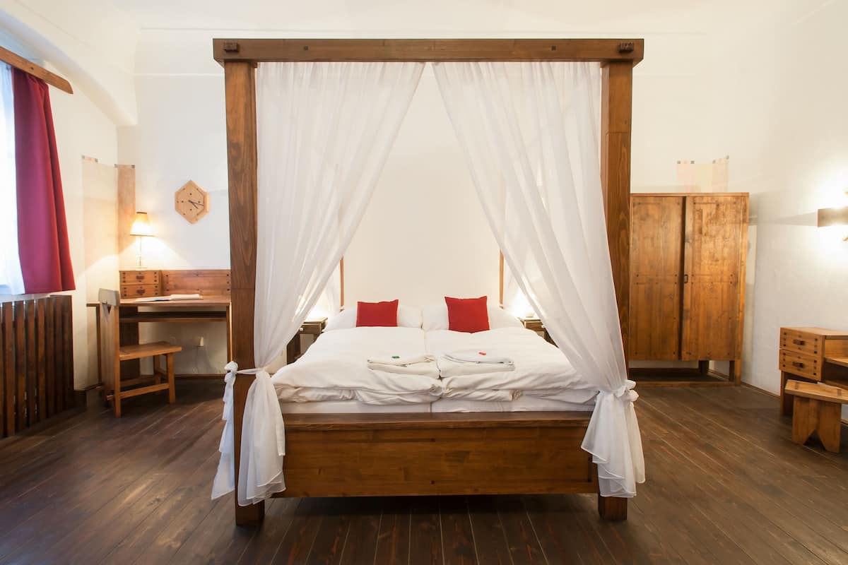 Uma cama de dossel com cortinas, roupa de cama branca, uma escrivaninha de madeira e um guarda-roupa de madeira num quarto com soalho de madeira.