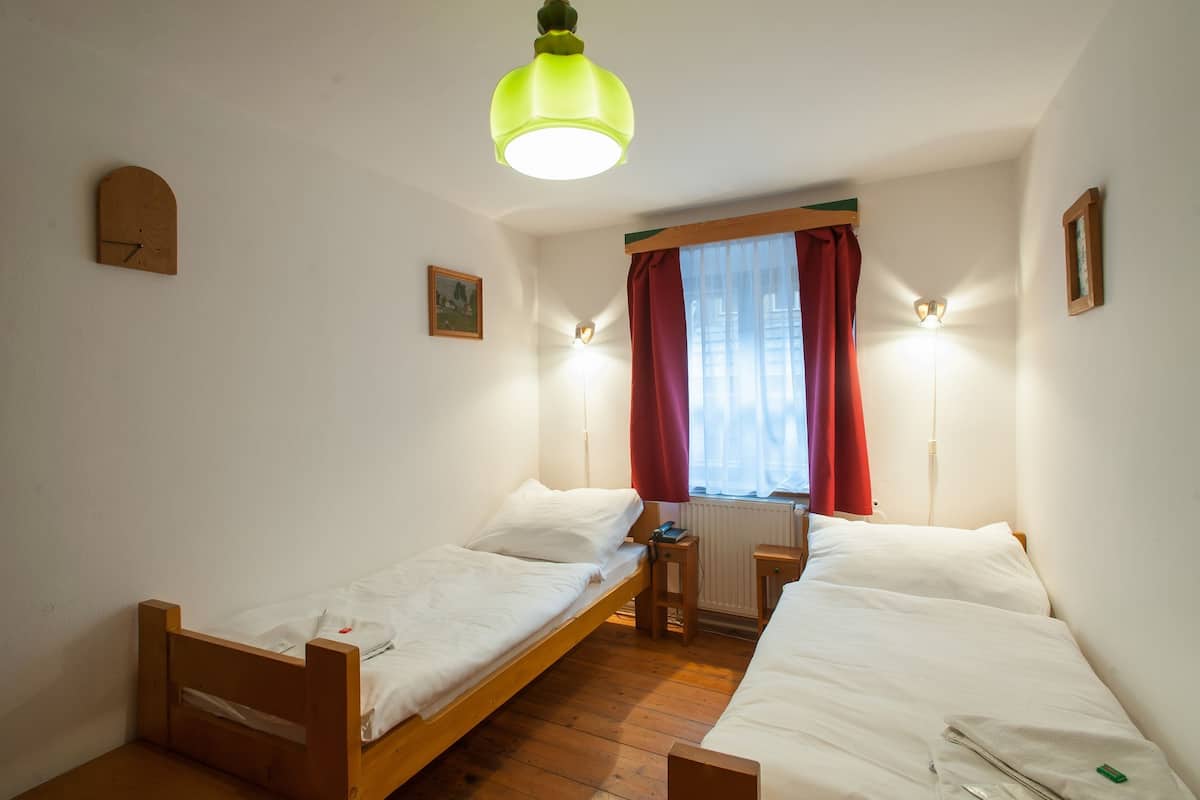 Quarto pequeno e limpo com duas camas de solteiro, cabeceira de madeira, um banquinho pequeno, janela com cortinas vermelhas e uma luminária pendente verde.
