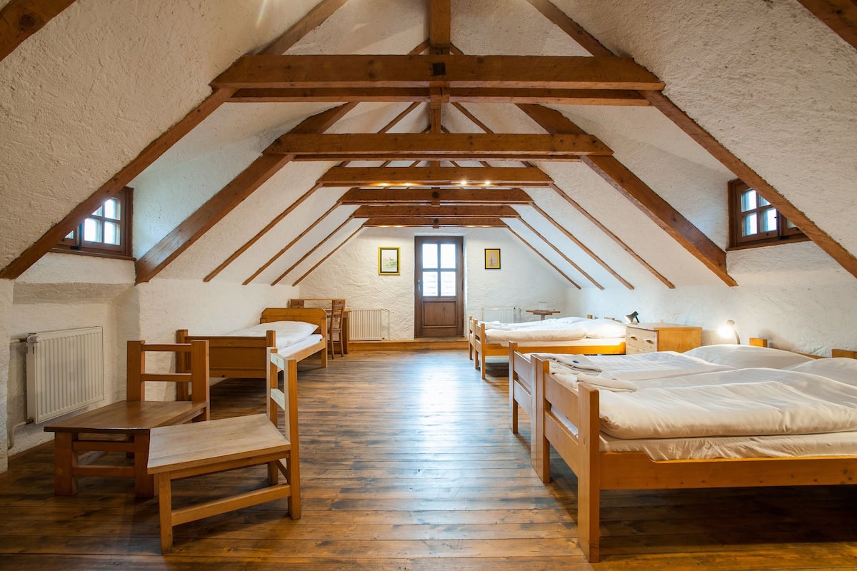 Quarto com vigas de madeira, piso de madeira e porta com duas janelas. Há camas e uma mesa com cadeiras.