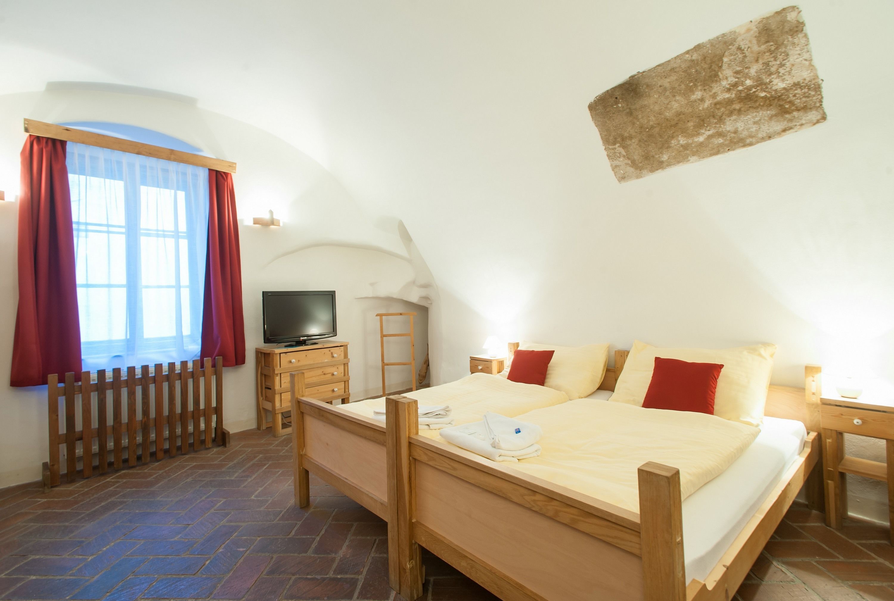 Quarto com cama de madeira, televisão, mesa de cabeceira de madeira e janela com cortinas vermelhas.