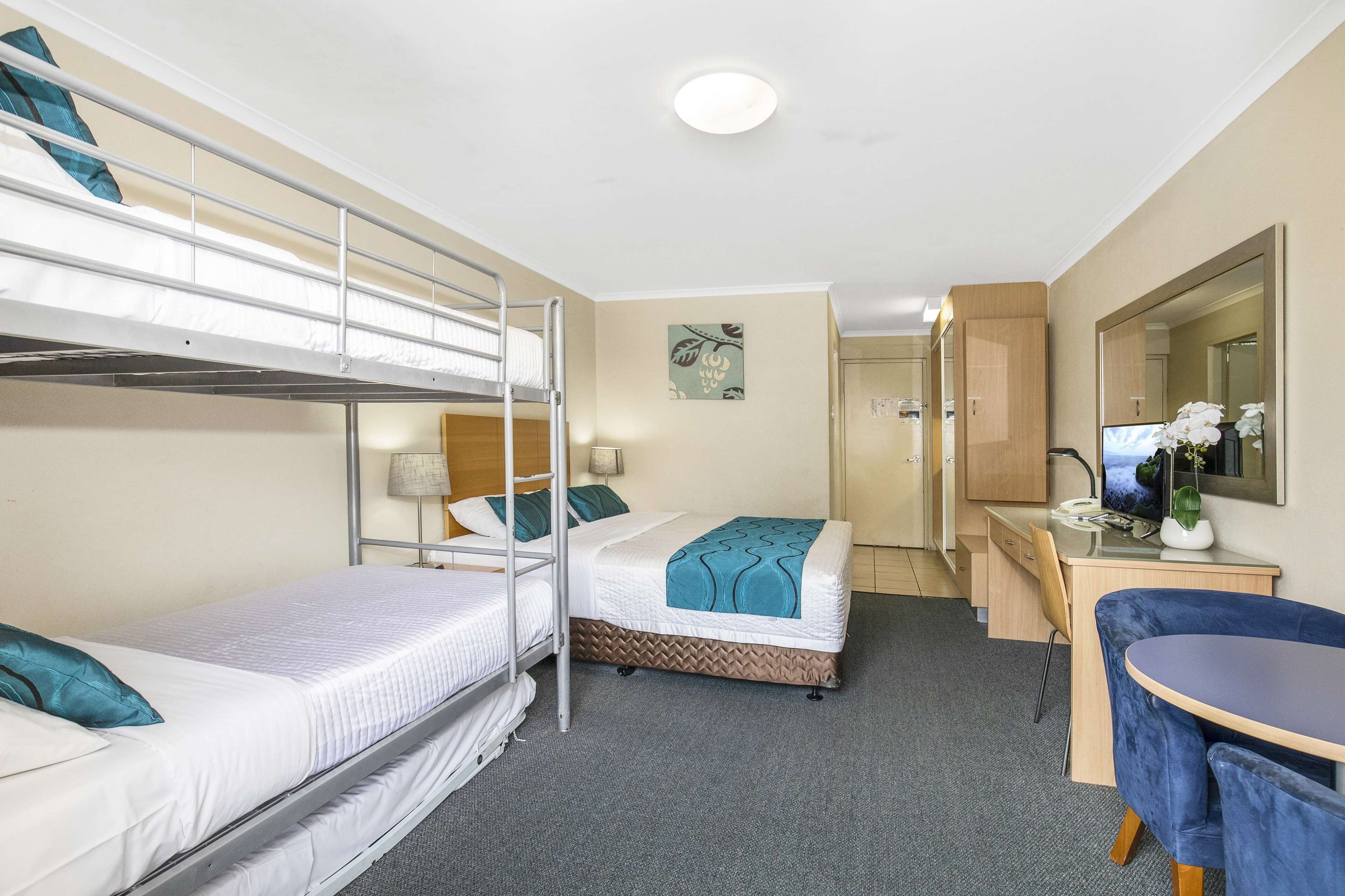 Quarto de hotel com beliches, uma escrivaninha, uma cadeira, televisão e espelho.