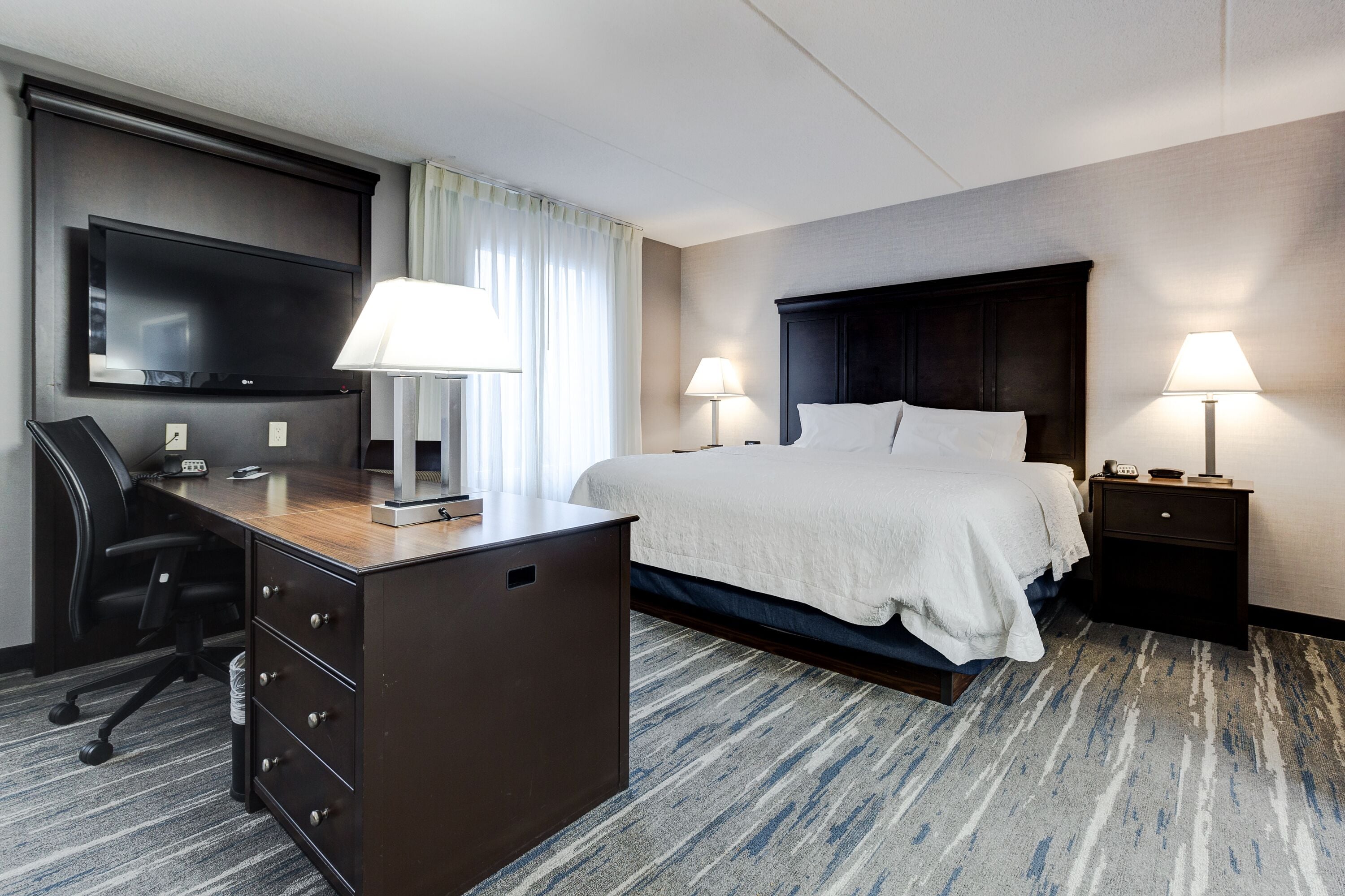 Quarto de hotel com cama, escrivaninha, cadeira, televisão e luminárias.