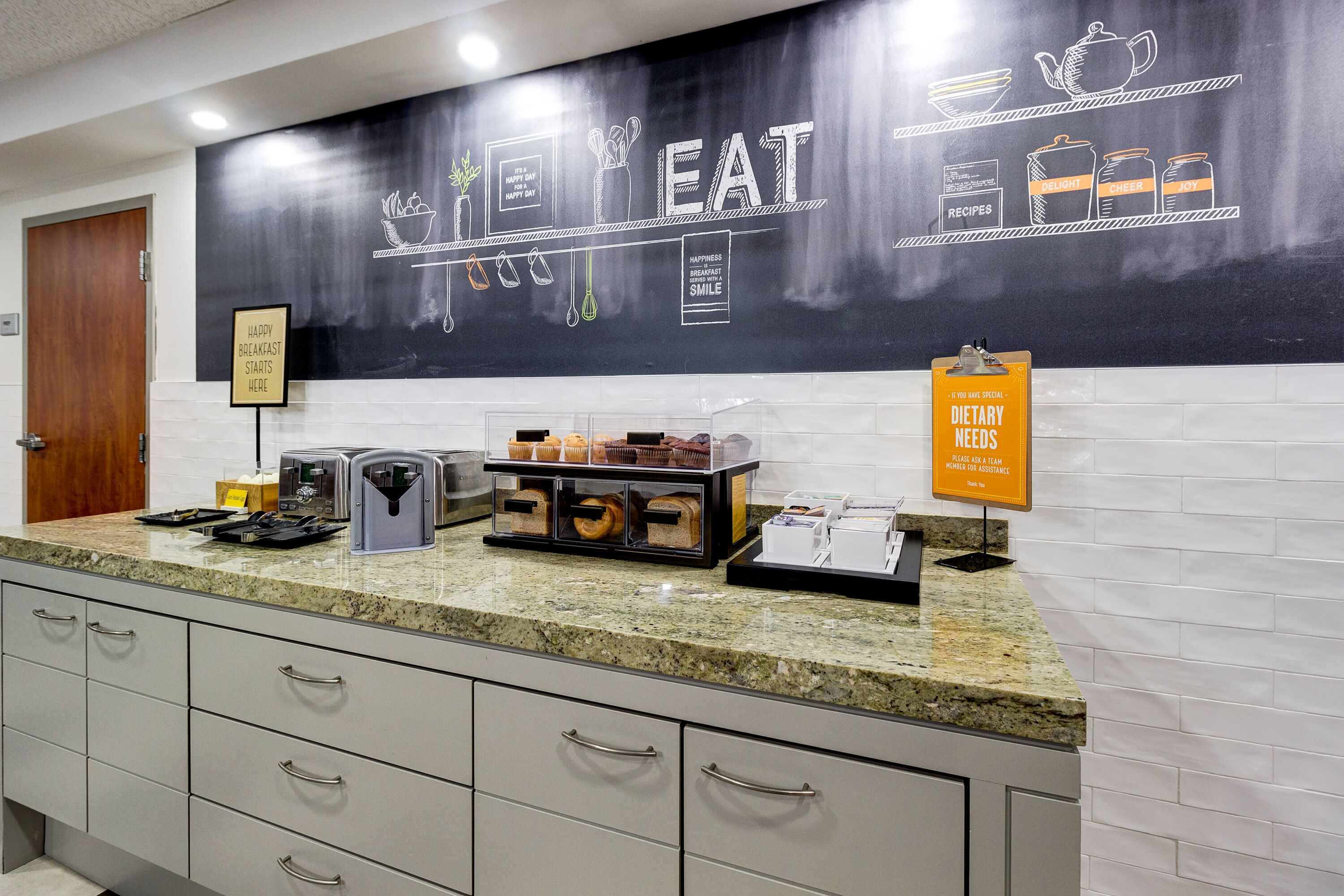 Espaço de refeições com parede de lousa exibindo a palavra "EAT" e diversos desenhos relacionados a alimentos. Há bandejas de comida, uma máquina de café e um aviso sobre necessidades alimentares.
