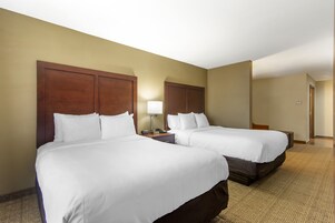 Quarto de hotel com duas camas, mesa de cabeceira com abajur e chão coberto de carpete.
