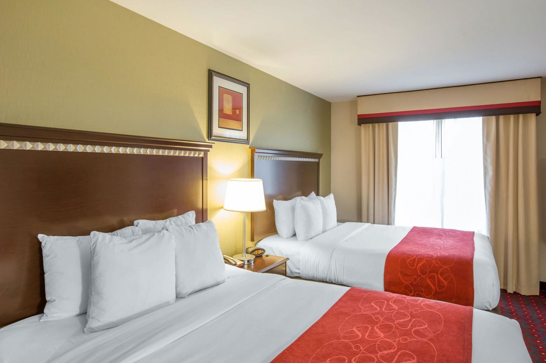 Quarto de hotel com duas camas, uma mesa de cabeceira, uma luminária, um quadro na parede e uma janela com cortinas.