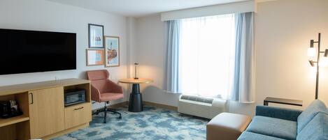 Quarto de hotel com cama, escrivaninha, cadeira, sofá, televisão, janela com cortinas e abajur.