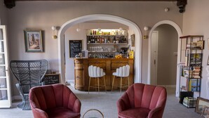 Uma sala de estar aconchegante com duas poltronas vermelhas, uma mesa de centro com revistas e uma tigela, um espaço bar com prateleiras de garrafas e copos, e uma estante com livros e uma moldura com foto.