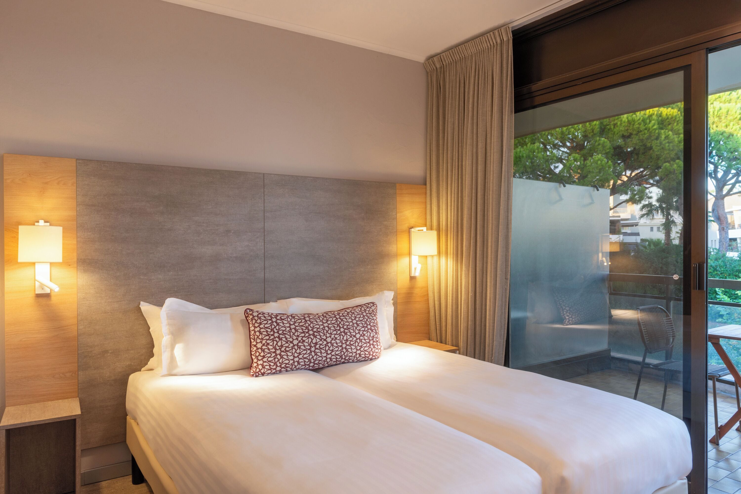 Uma cama de casal com lençóis brancos e almofada estampada, com duas luminárias fixadas na parede de cada lado.