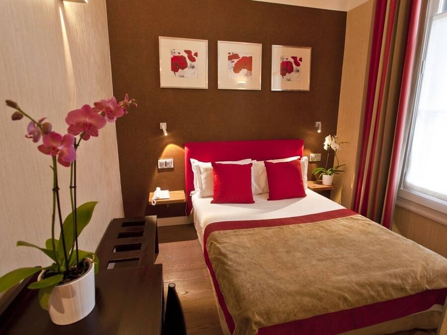 Quarto de hotel com uma cama grande, roupa de cama vermelha e branca, mesa de cabeceira com abajur e uma orquídea em vaso.