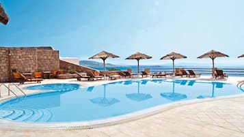 Uma área de piscina com espreguiçadeiras e guarda-sóis, uma parede de pedra e vista para o mar.