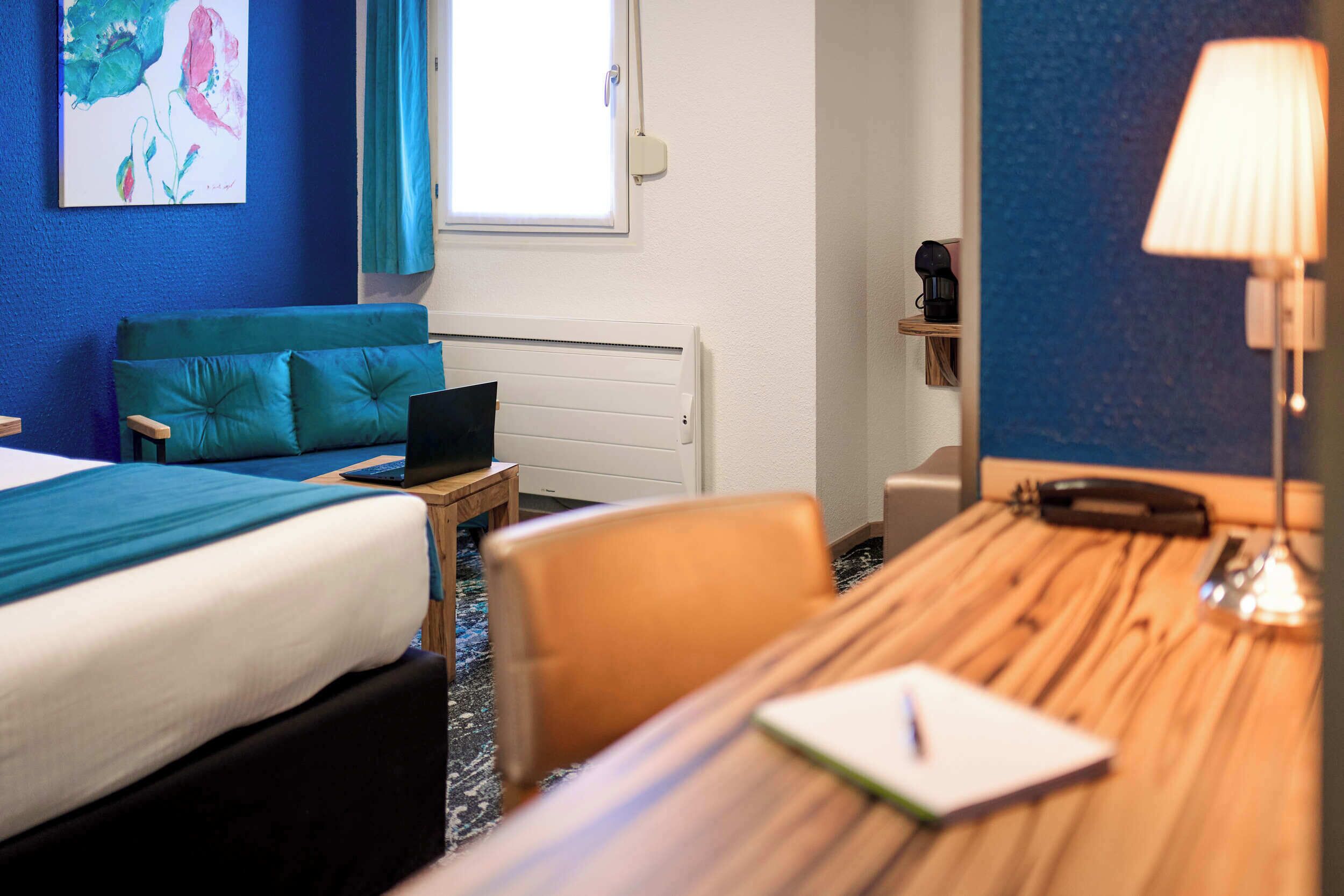 Quarto de hotel com cama, escrivaninha, cadeira, luminária, laptop e um quadro na parede.