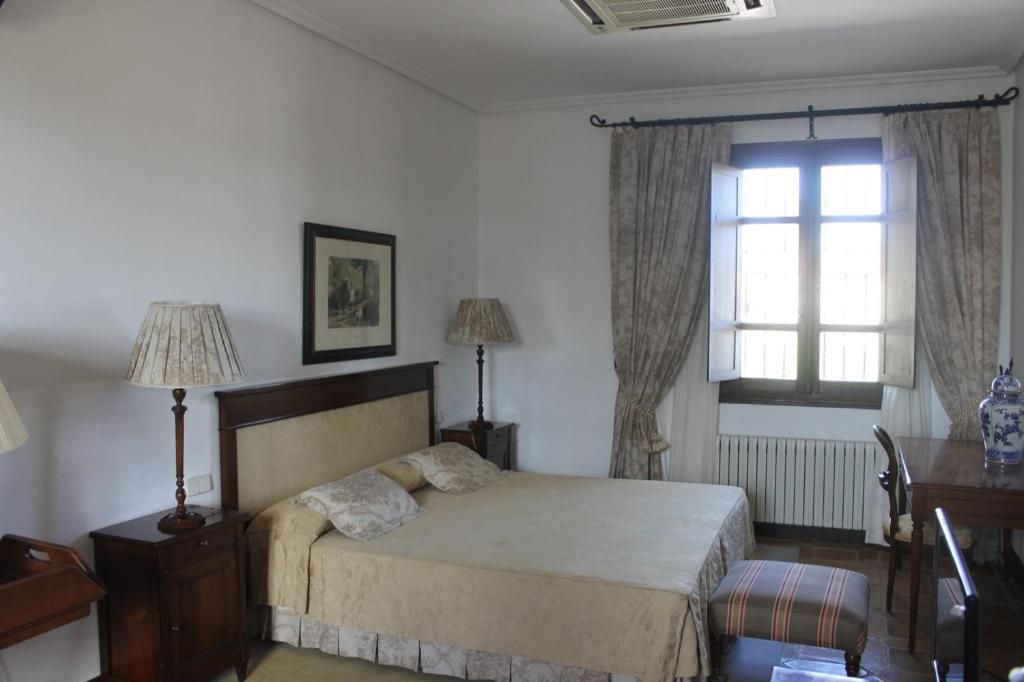 Quarto com cama, duas mesinhas de cabeceira, janela com cortinas, radiador e uma pequena mesa com um vaso.