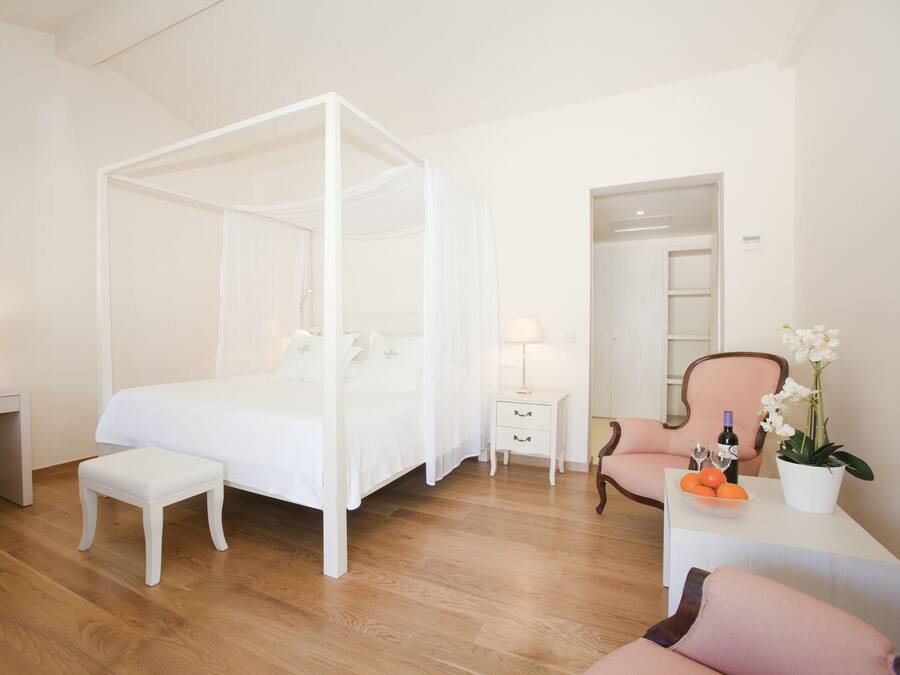 Quarto com cama de dossel, mesa de cabeceira com abajur, cadeira, escrivaninha com abajur e uma pequena mesa com garrafa e flores.